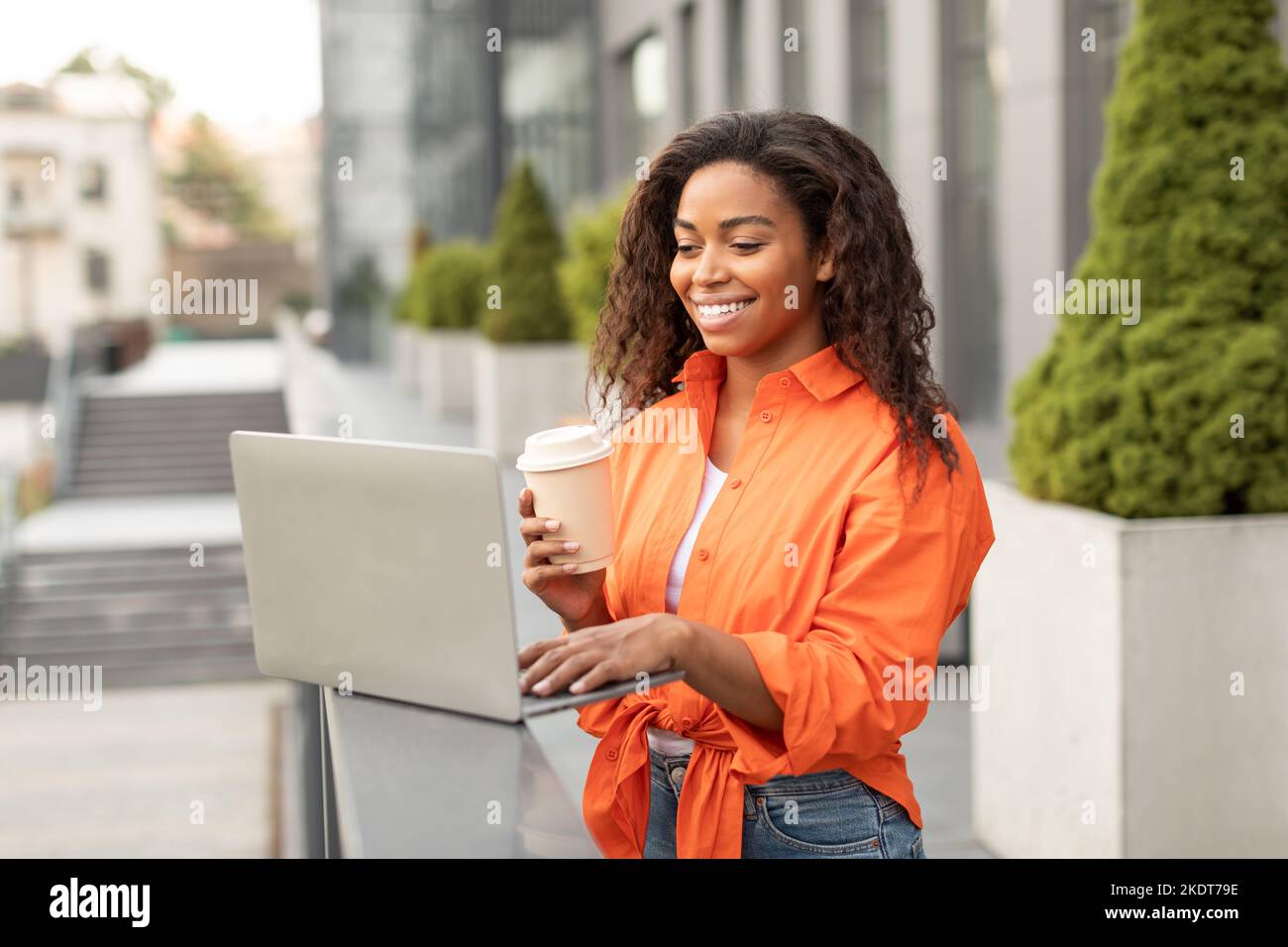 Smiling young black lady with cup of coffee to go chatting on computer ...