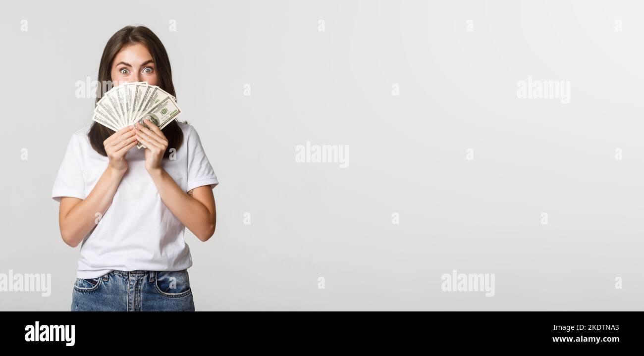 Excited pretty young woman holding money over face, standing white ...