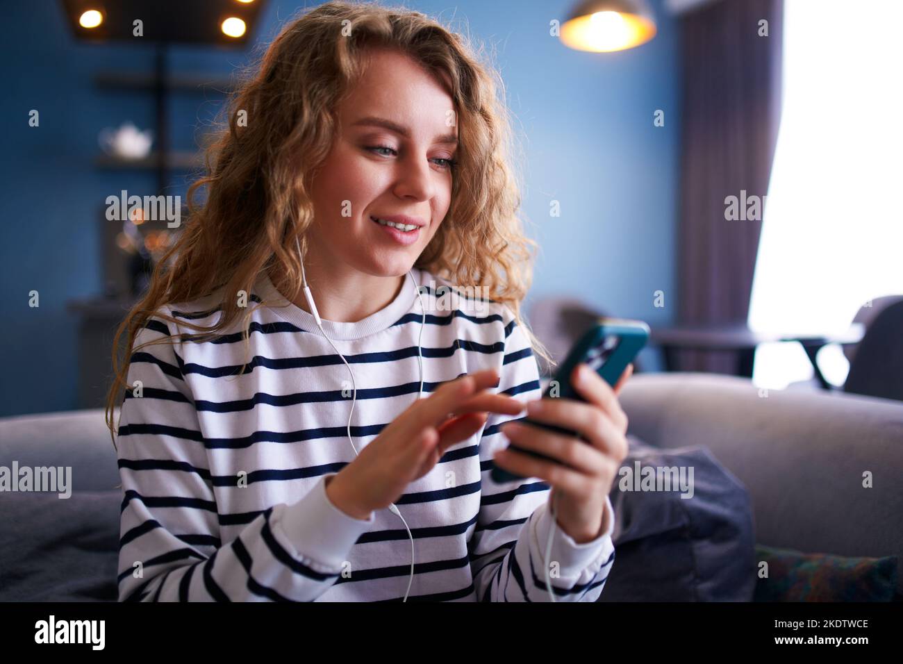 Woman scrolling smartphone, web surfing, sitting on sofa at stylish living room at night. Female ...