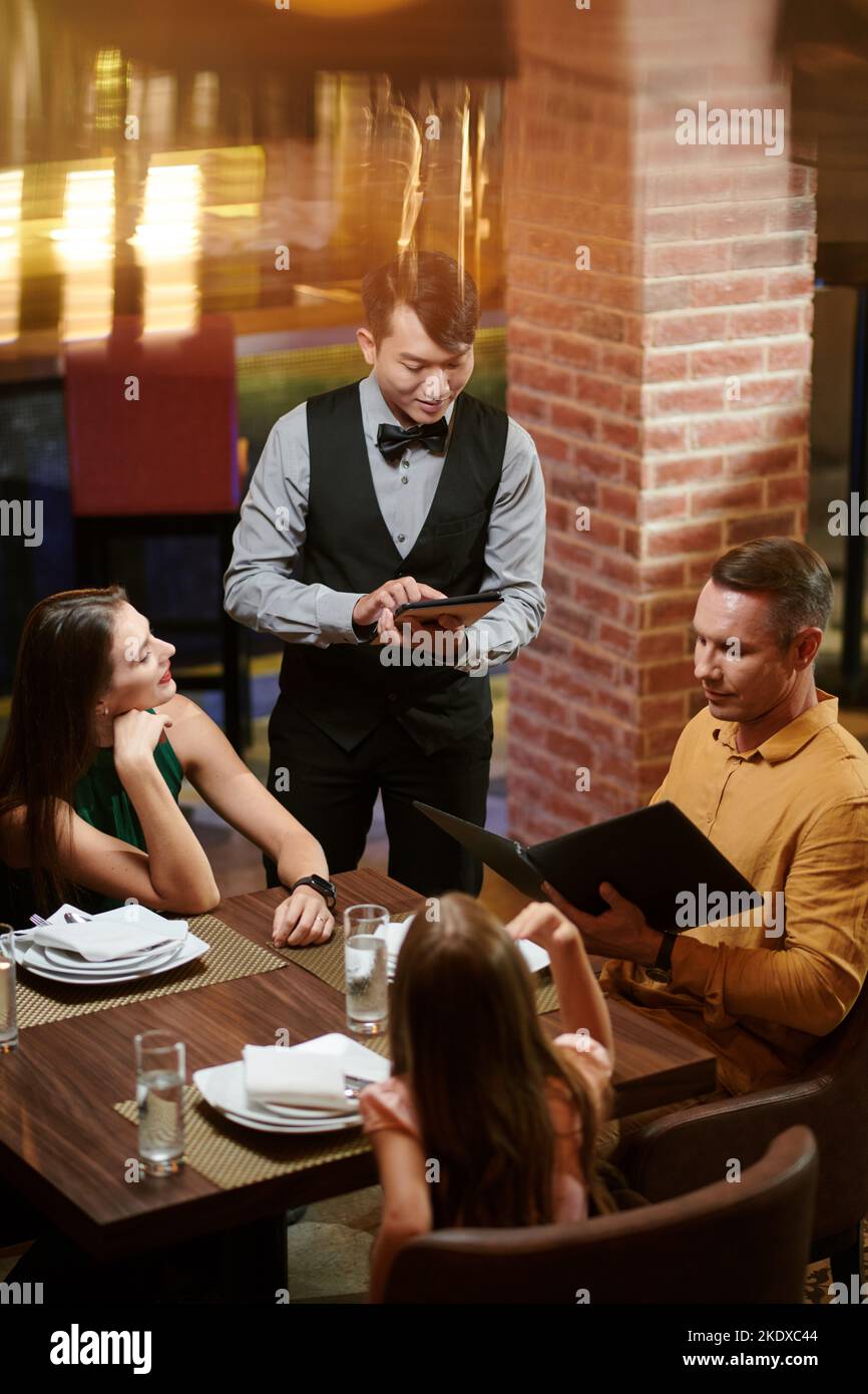Smiling waiter taking notes on tablet computer when family ordering ...