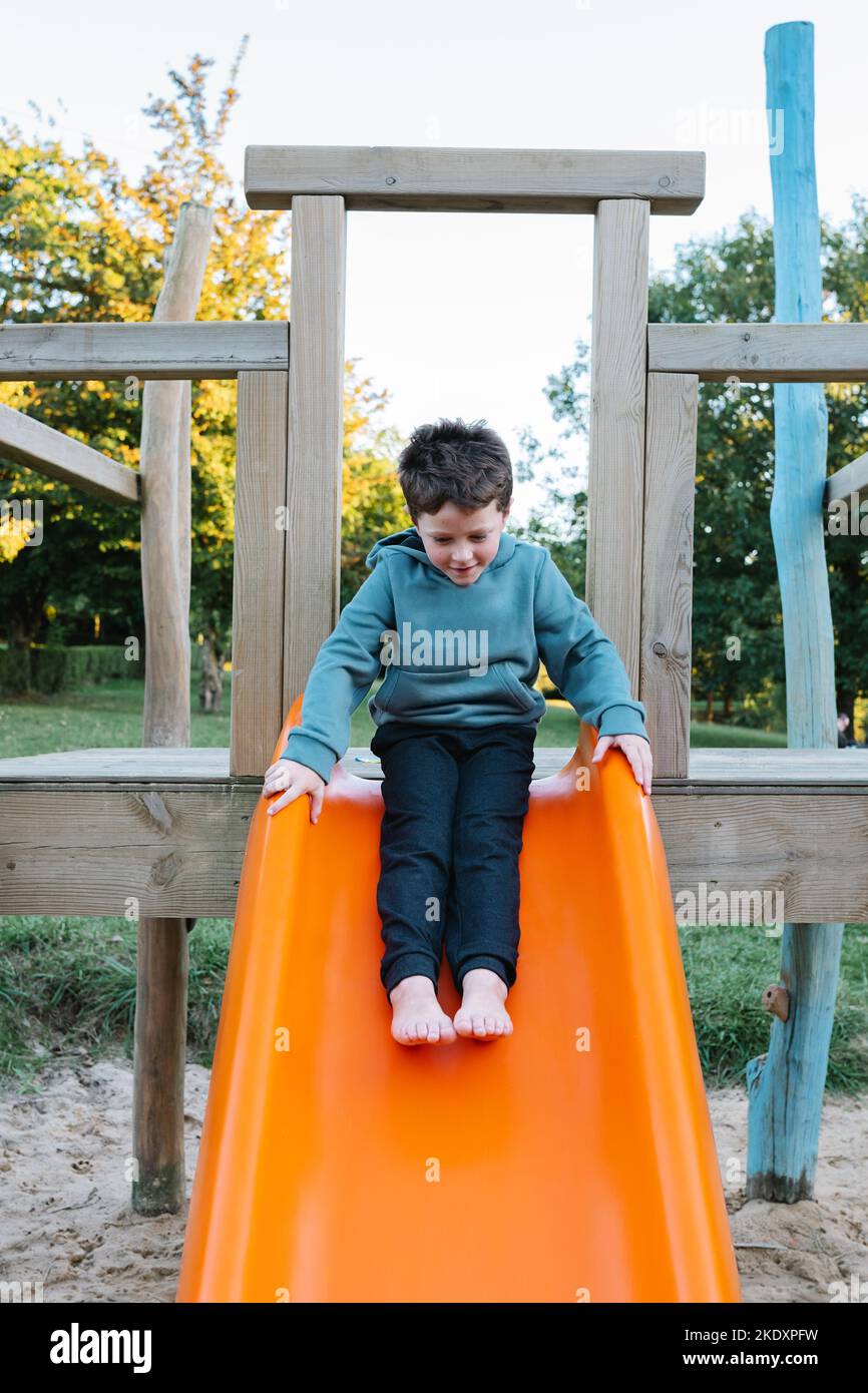 Full body barefoot boy sitting on orange slide while playing on ...