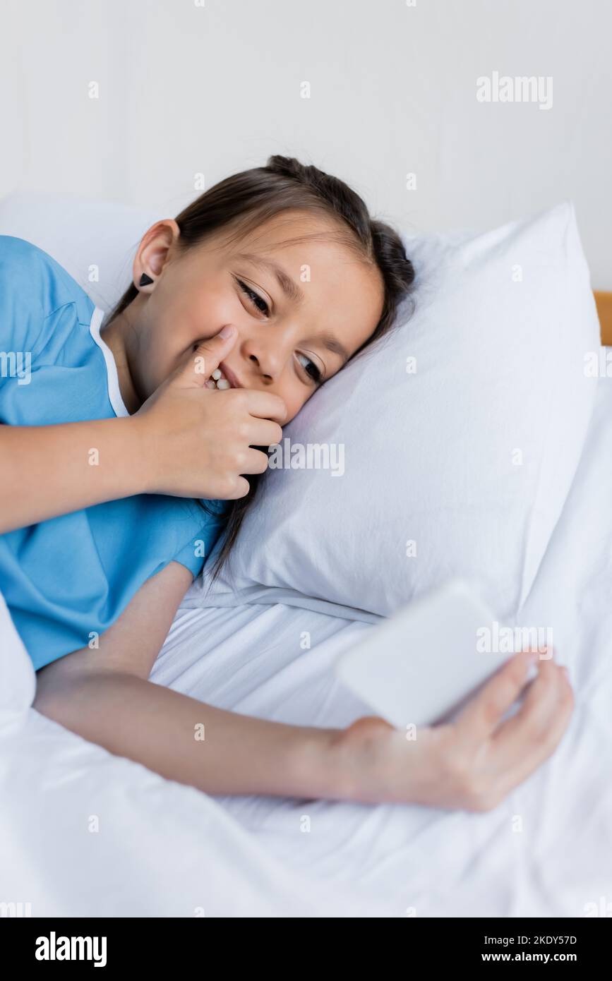 Cheerful kid using blurred mobile phone while lying on bed in hospital ...