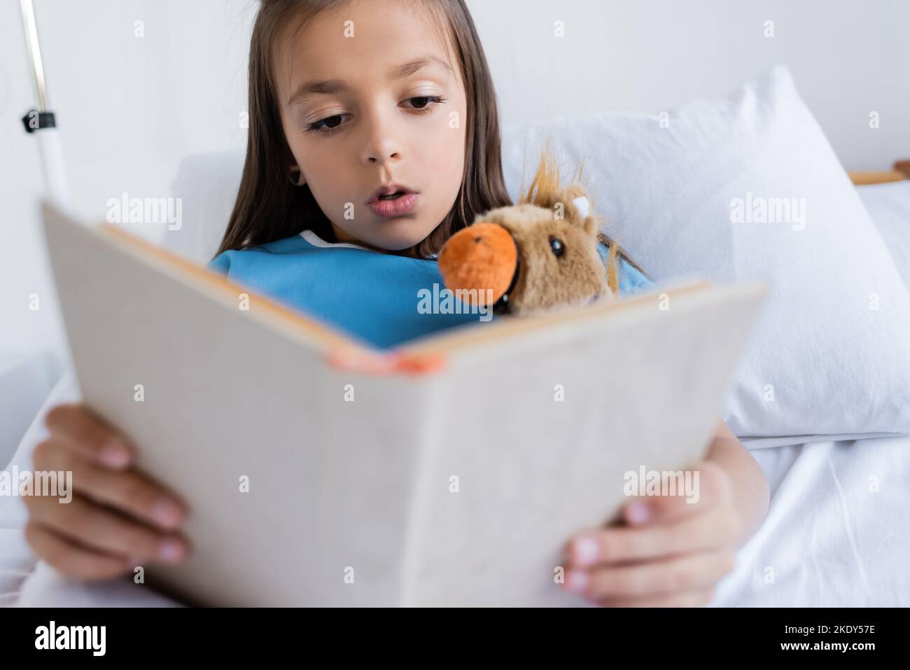 Kid in patient gown reading book near toy on hospital bed,stock image ...