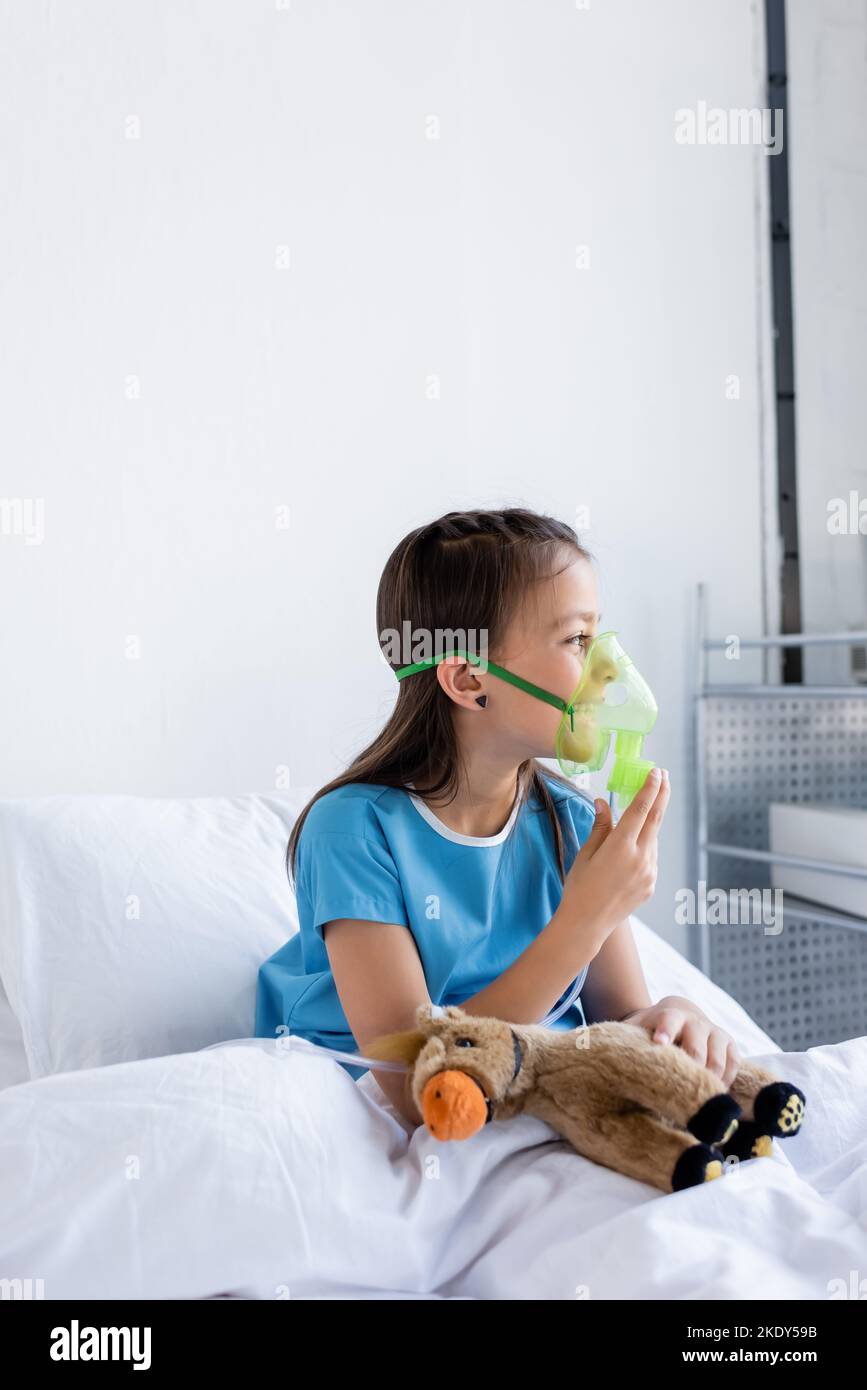 Side view of child holding oxygen mask and toy on bed in hospital ward ...