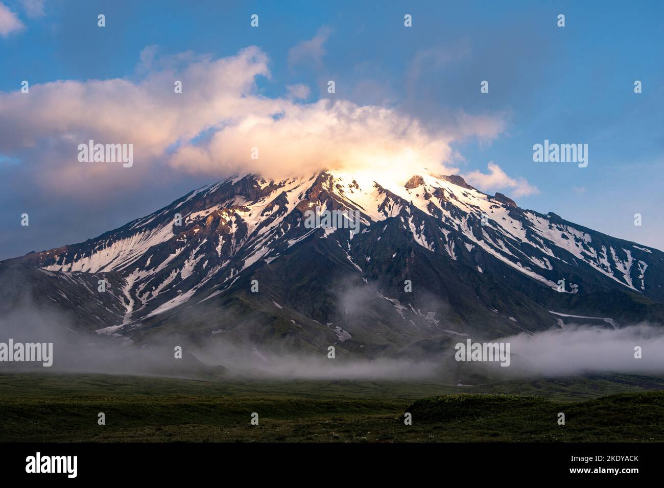 Smoking volcano covered with snow, green valley, blue sky with clouds ...