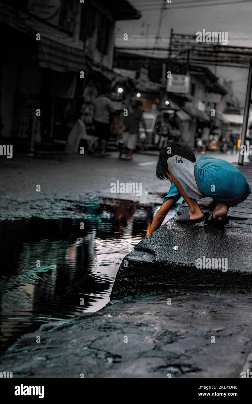 A vertical shot of a poor child playing with water from the rain in ...