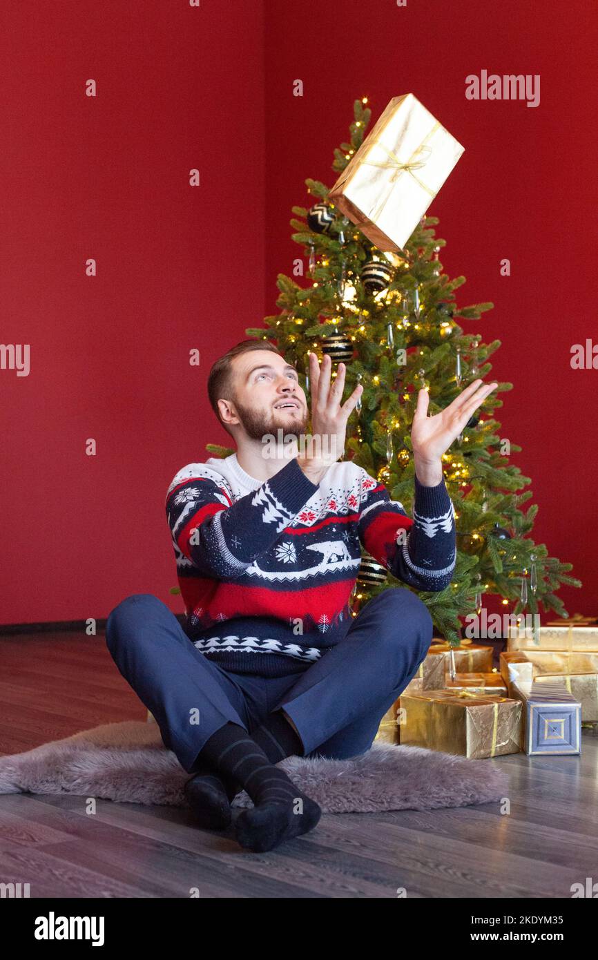 Cute man in sweater sits by nice Christmas tree and throws up his gold ...