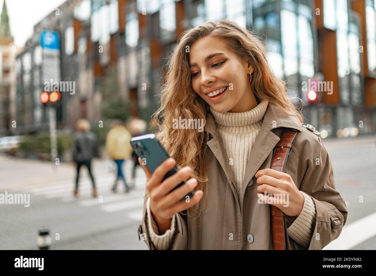 Smiling curly woman wearing warm coat walking down the street and using ...