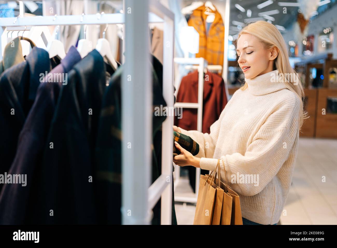 Focused elegant blonde young woman choosing coat in clothing store ...