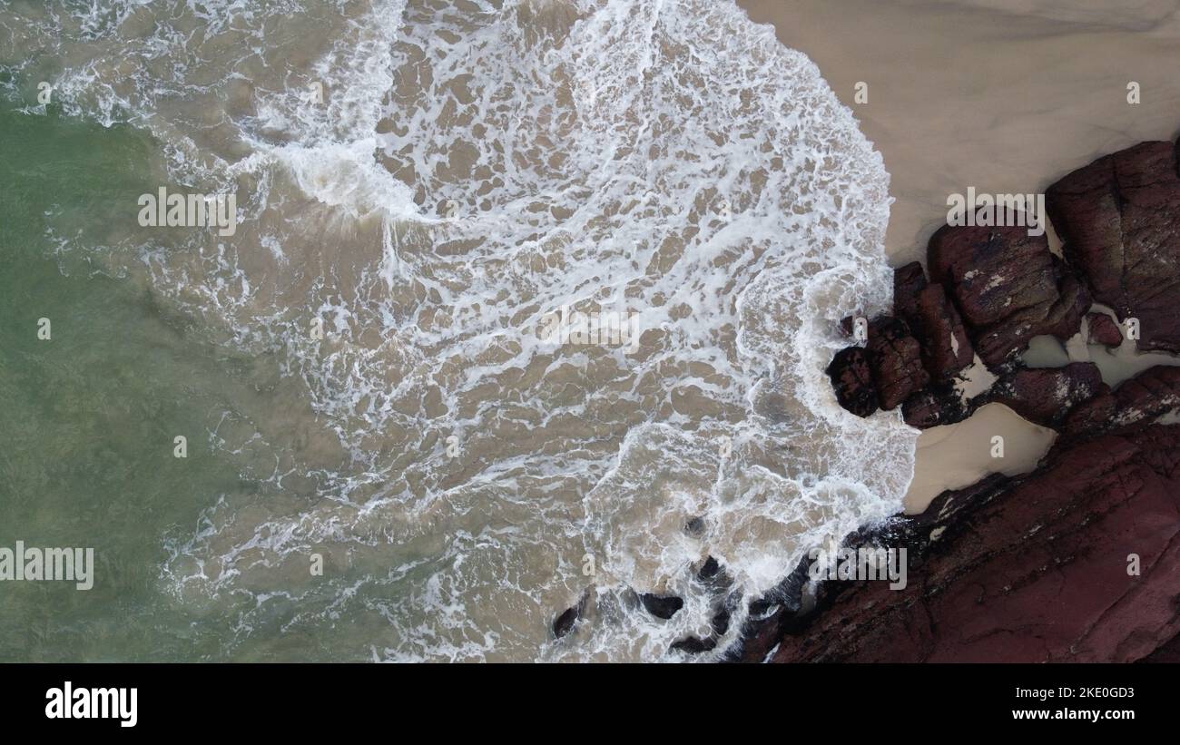 A top view of a wavy sea washing over the sandy seashore and the rocky ...