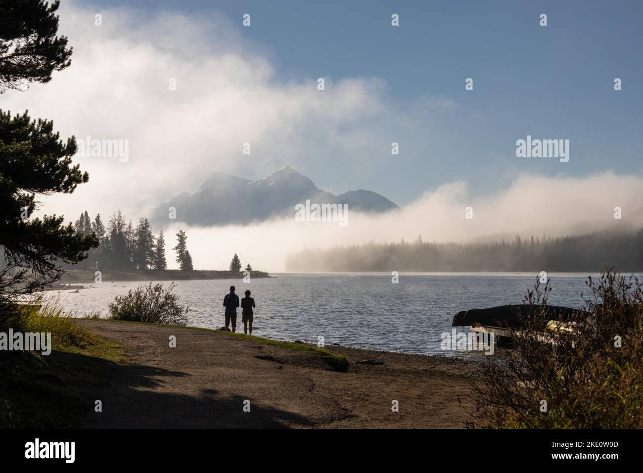 A silhouette of two people standing on a lake coast in foggy weather ...