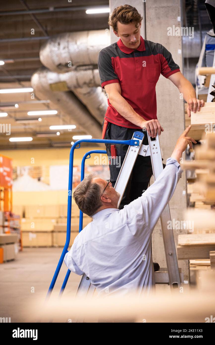 Male customer showing plank to salesman standing on ladder at hardware ...