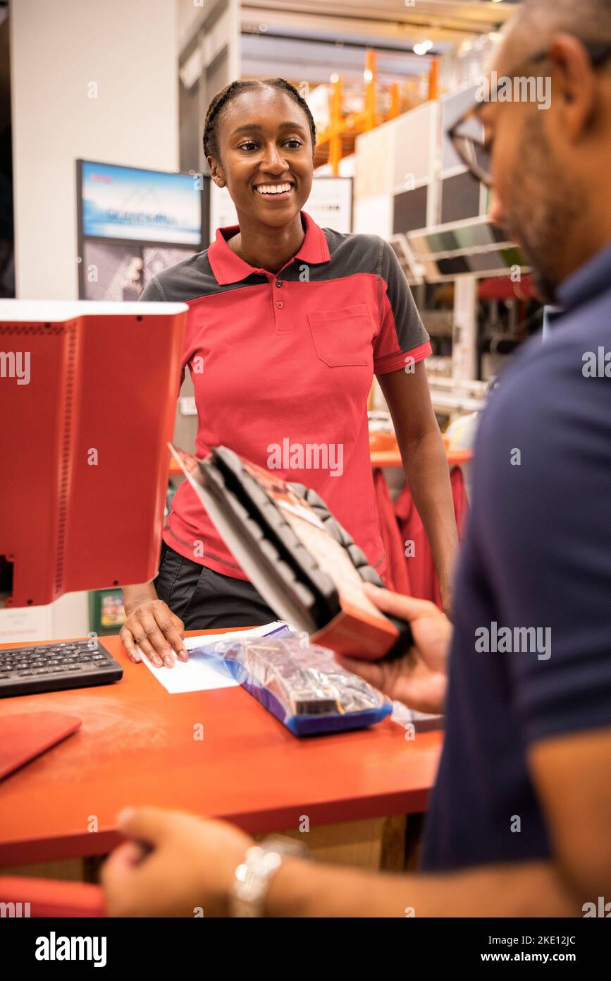 Smiling female cashier looking at male customer holding merchandise at ...