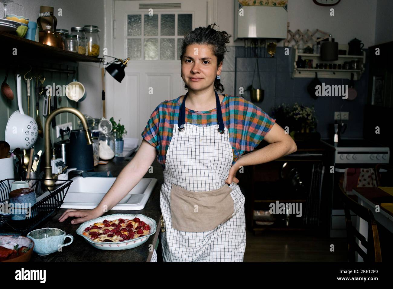 Portrait of woman with hand on hip standing by pie on kitchen counter ...