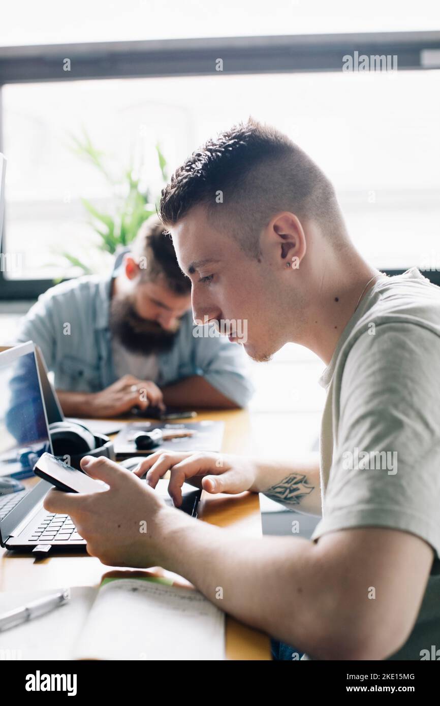 Male computer programmer using smart phone by laptop on desk in startup ...