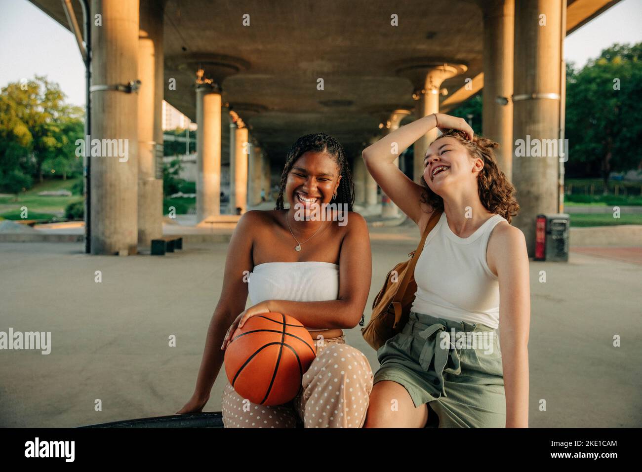 Cheerful teenage girl with basketball sitting by female friend under ...