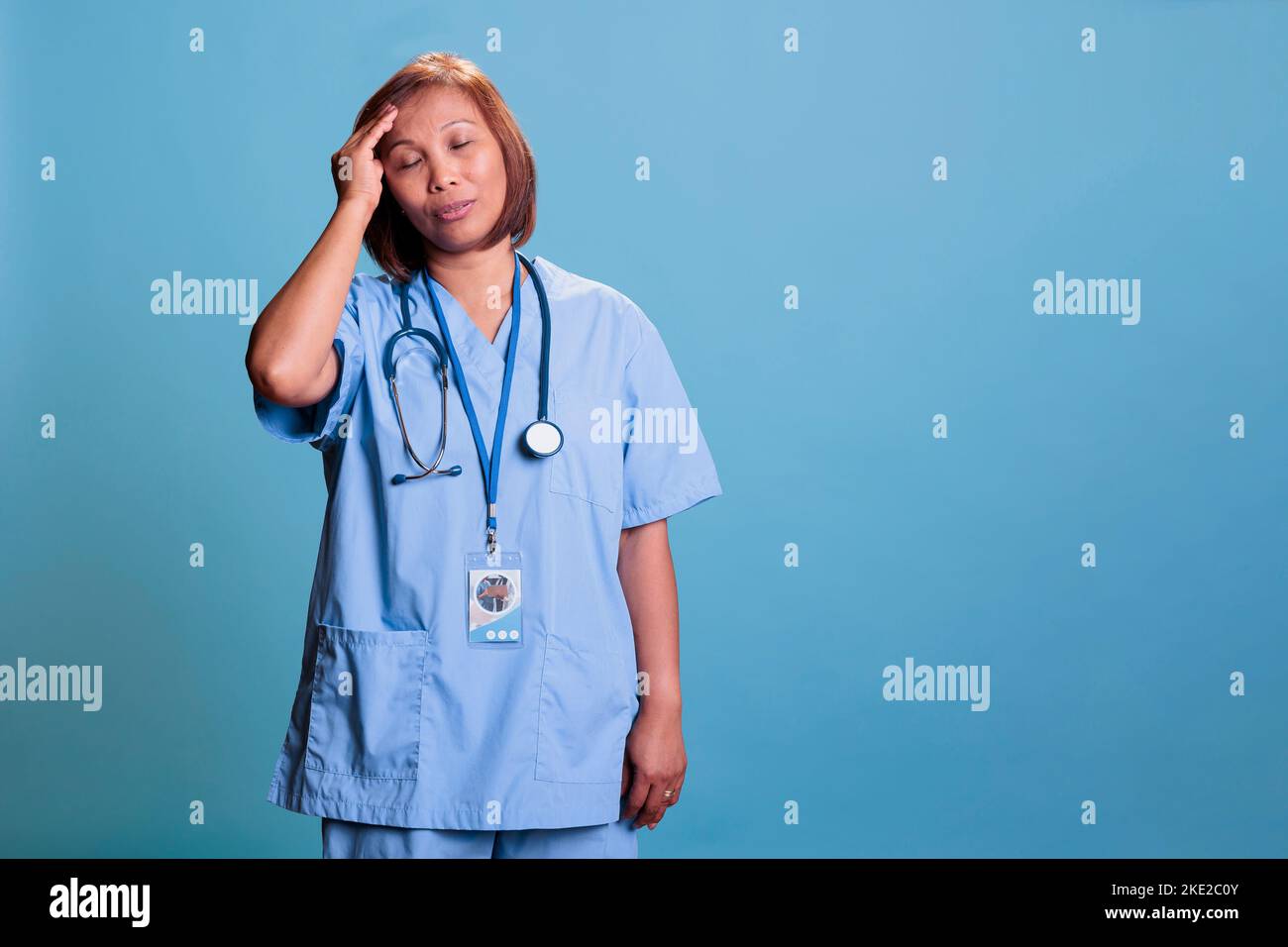 Tired asian assistant standing in studio with blue background suffering ...