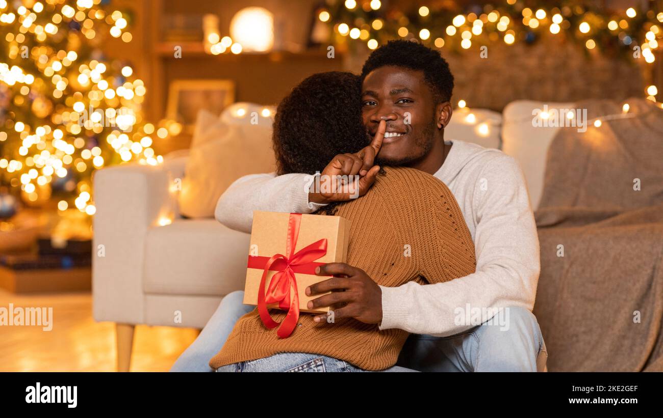 Happy young black wife hug husband with gift box, man show shh sign in ...