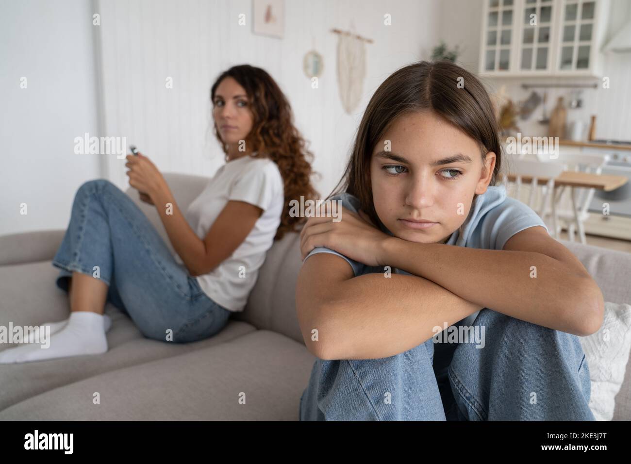 Casual worried woman sits on sofa behind sad teen girl leans on knees ...