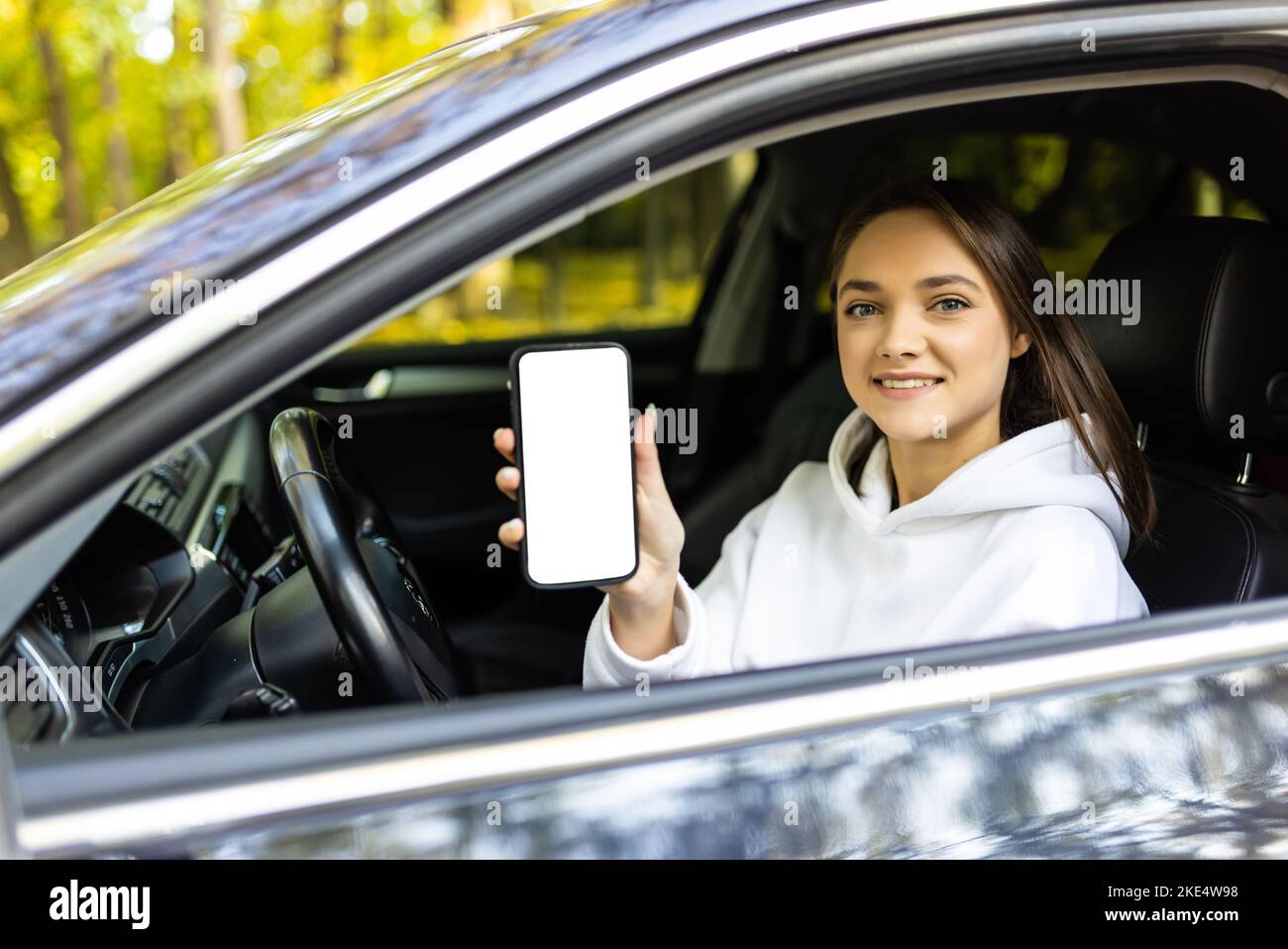 A young beautiful woman driving a car, she points to blank phone screen ...