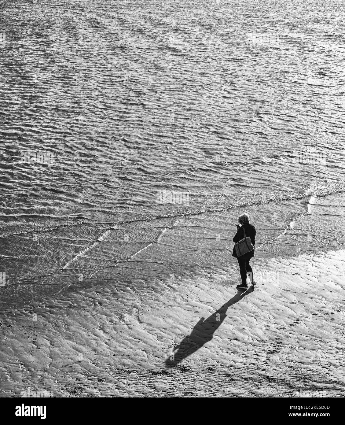 A solo woman stands on a beach by the incoming sea. Her shadow falls on ...