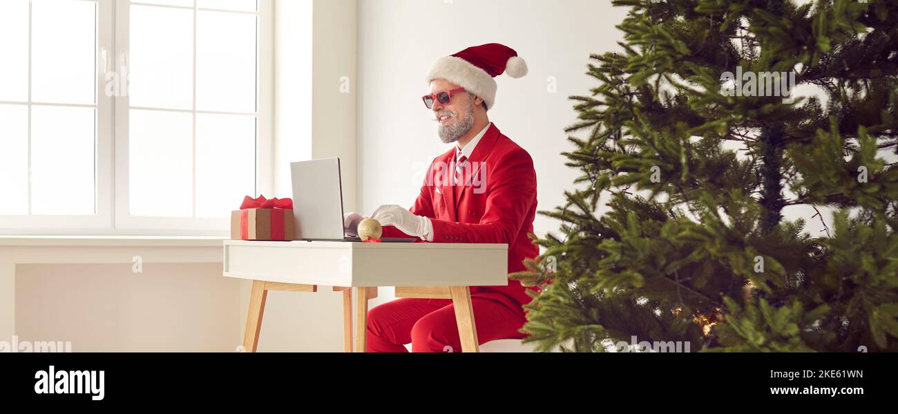 Happy grey-haired bearded man in red suit, Santa cap sits at desk ...