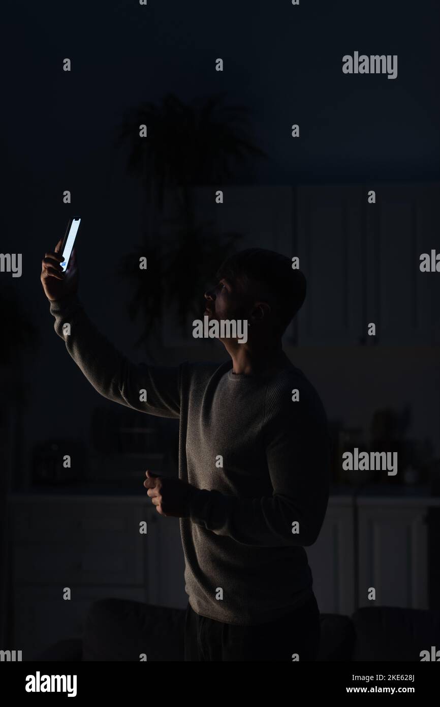 man standing in dark kitchen with smartphone in raised hand and ...