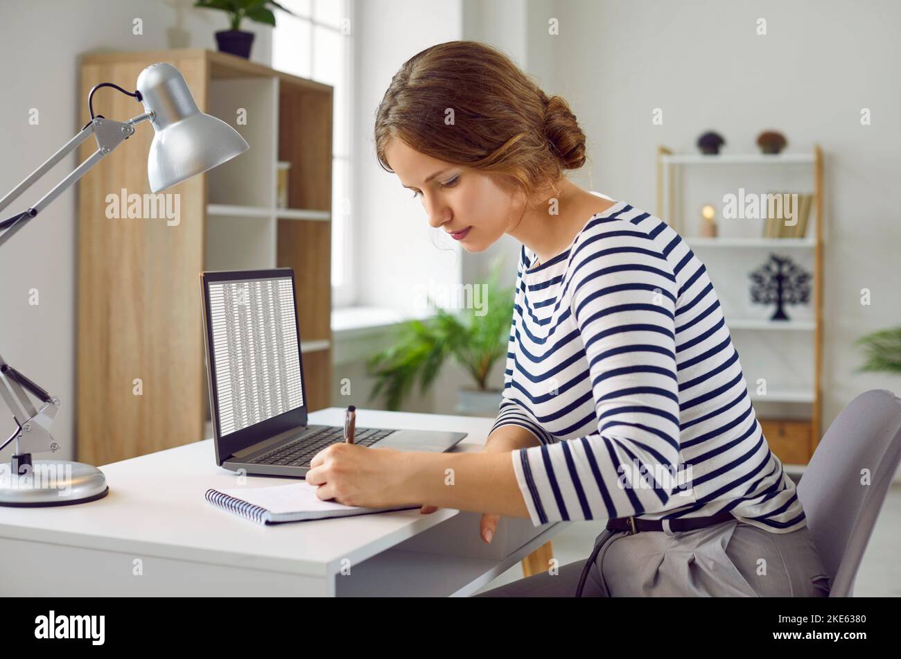 Left-handed woman makes notes in her notebook while working on laptop ...