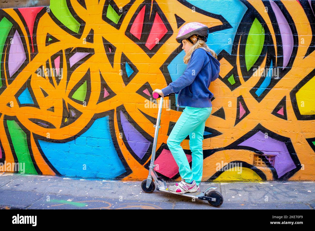 A side view of a young girl with scooter, standing next to a graffiti ...