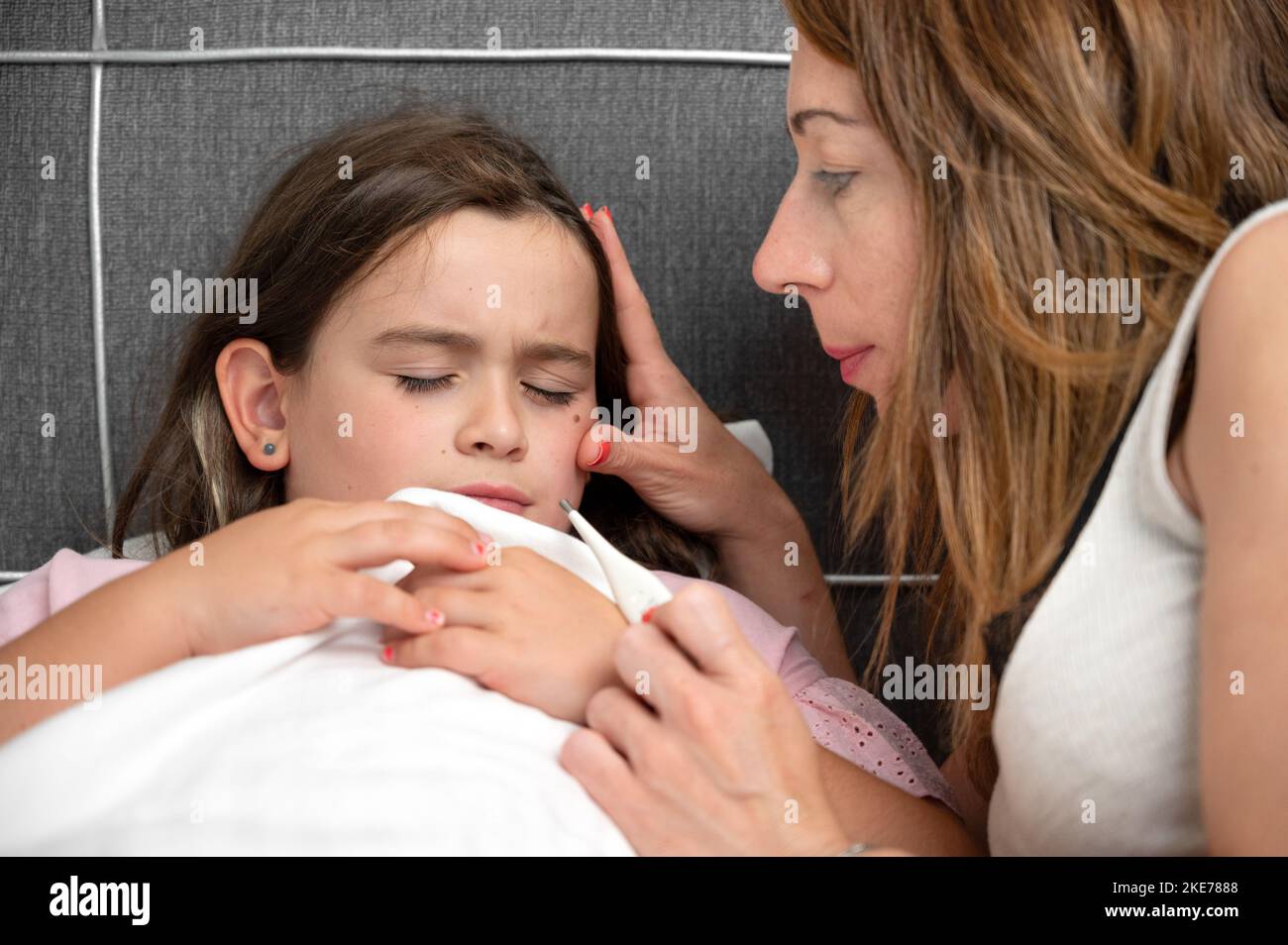 Mother measuring temperature of her ill daughter. Sick child with high fever laying in bed and ...