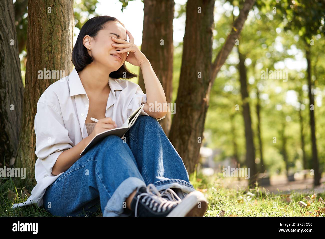 Sad girl crying while writing in her diary, sitting alone under tree in ...