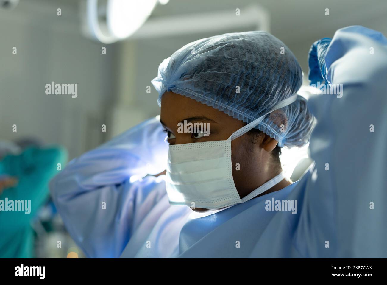 Biracial female surgeon in surgical cap and gown putting on mask in ...