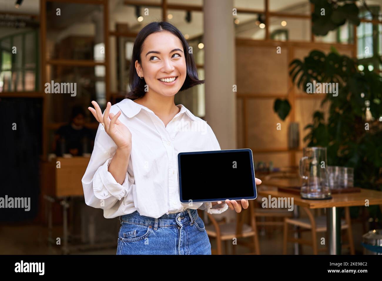 Friendly smiling asian woman, cafe manager, showing tablet screen and ...