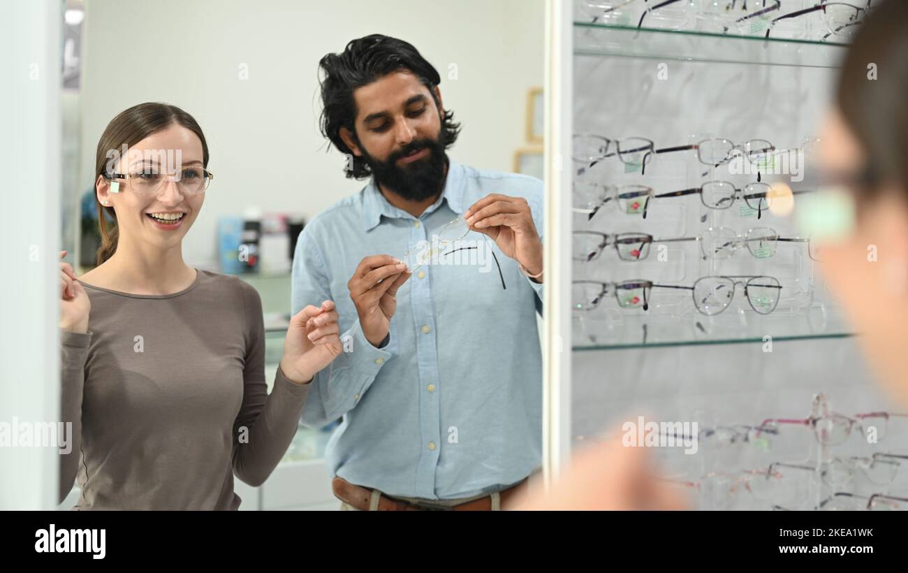 Happy young woman trying on spectacles and looking in the mirror at ...