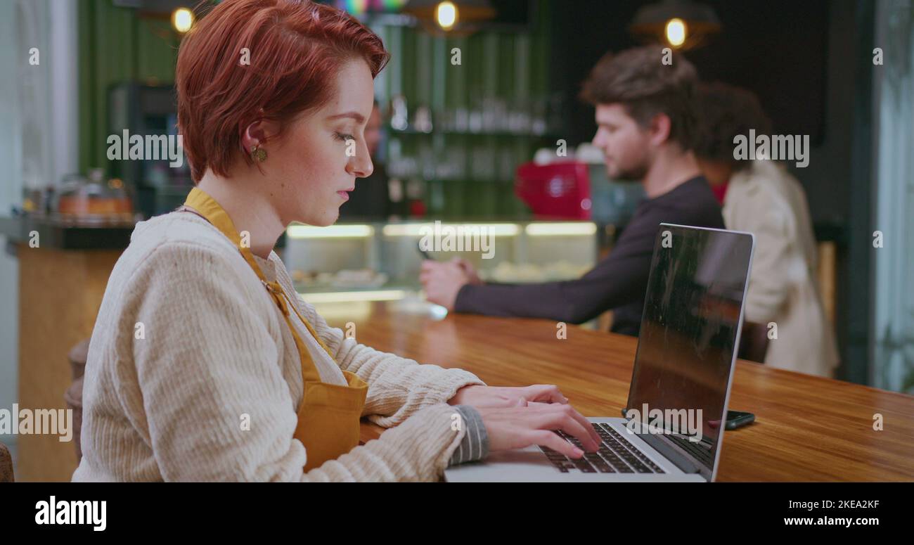 One young female barista using laptop seated at coffee shop staring at ...
