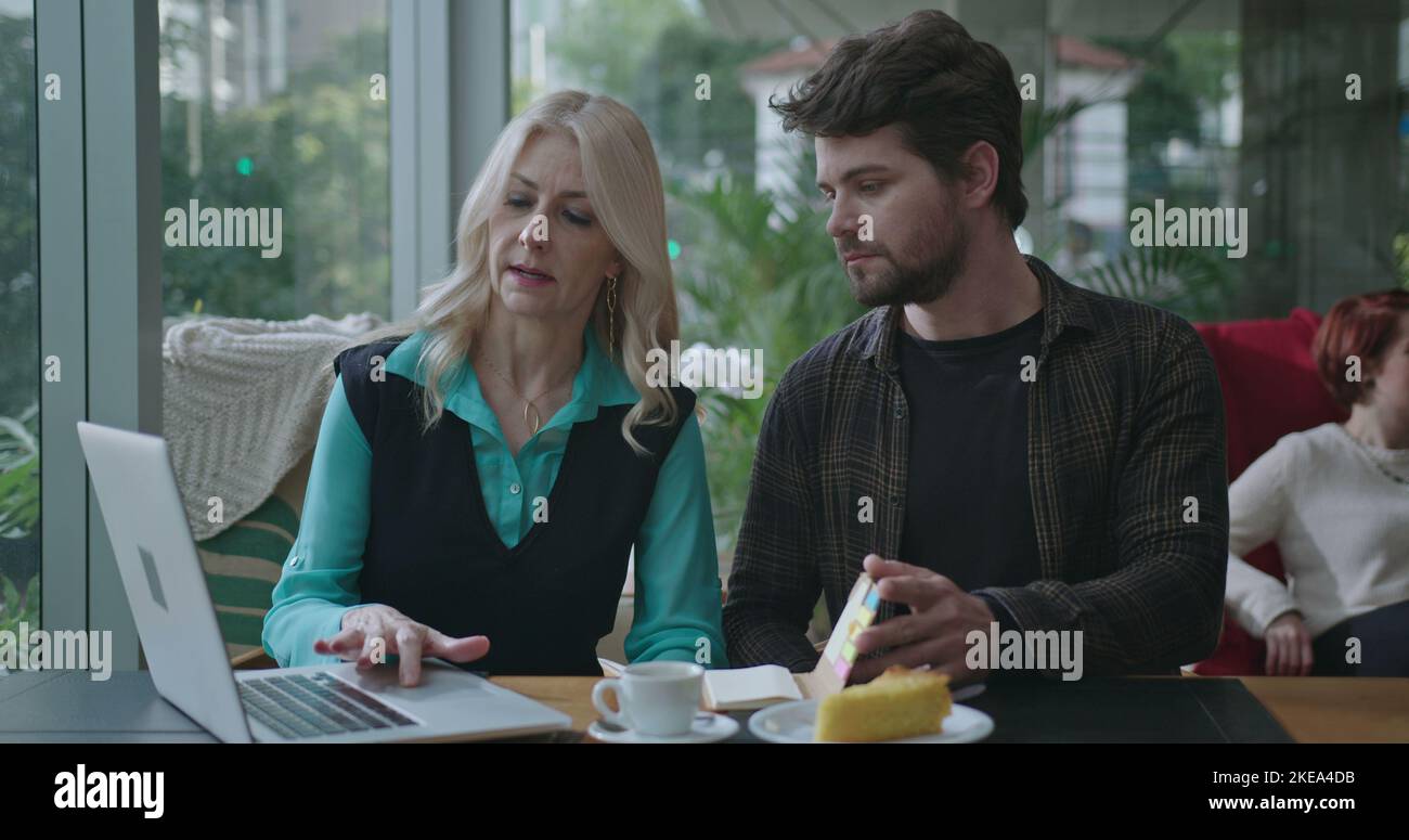 Senior female leader coaching male employee seated at coffee shop. Mature woman boss explaining ...