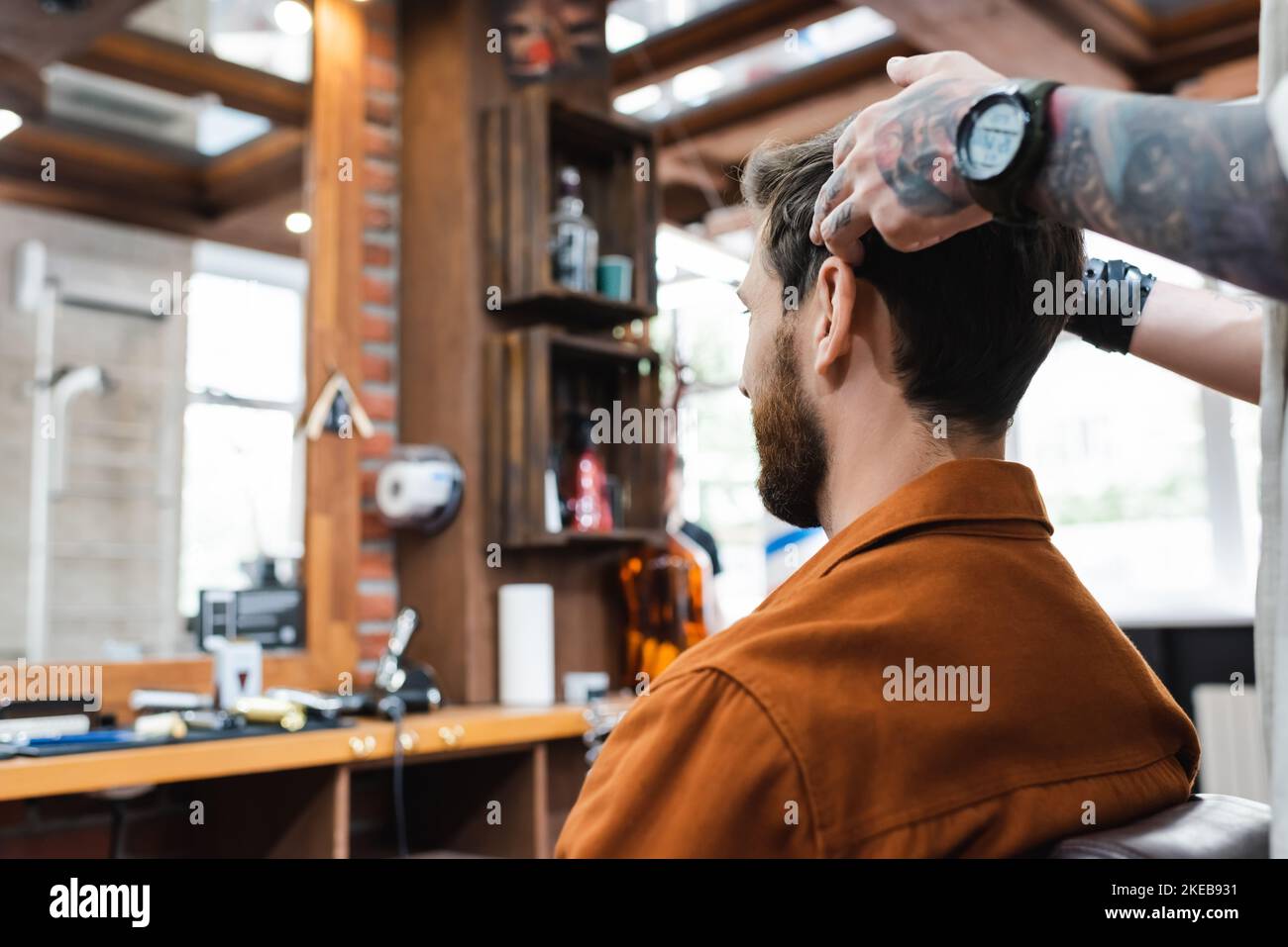Brunette Man Sitting In Barbershop Near Blurred Mirror While Hairdresser Touching His Hairstock