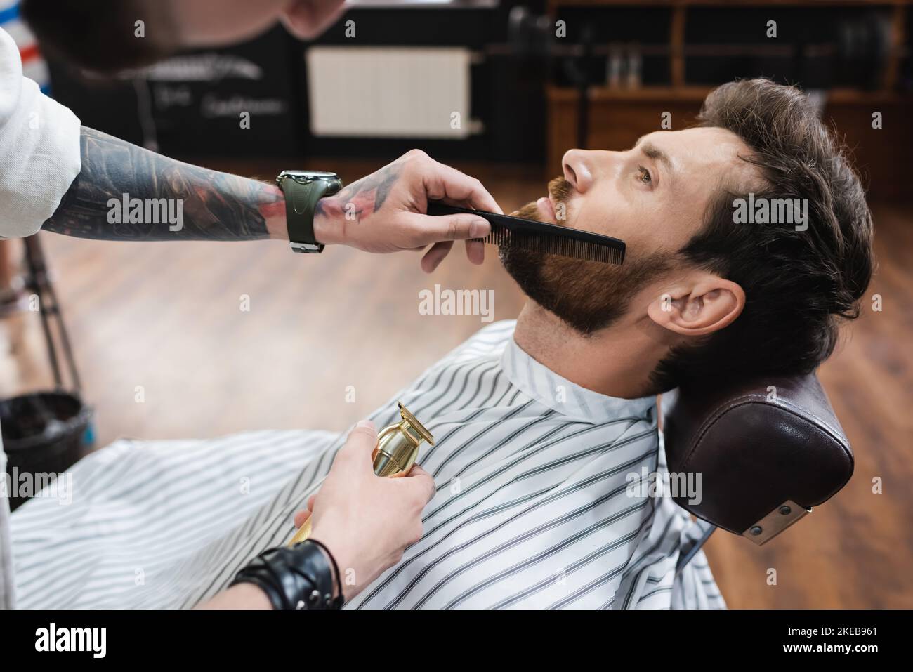 tattooed barber holding hair clipper and combing beard of man in ...