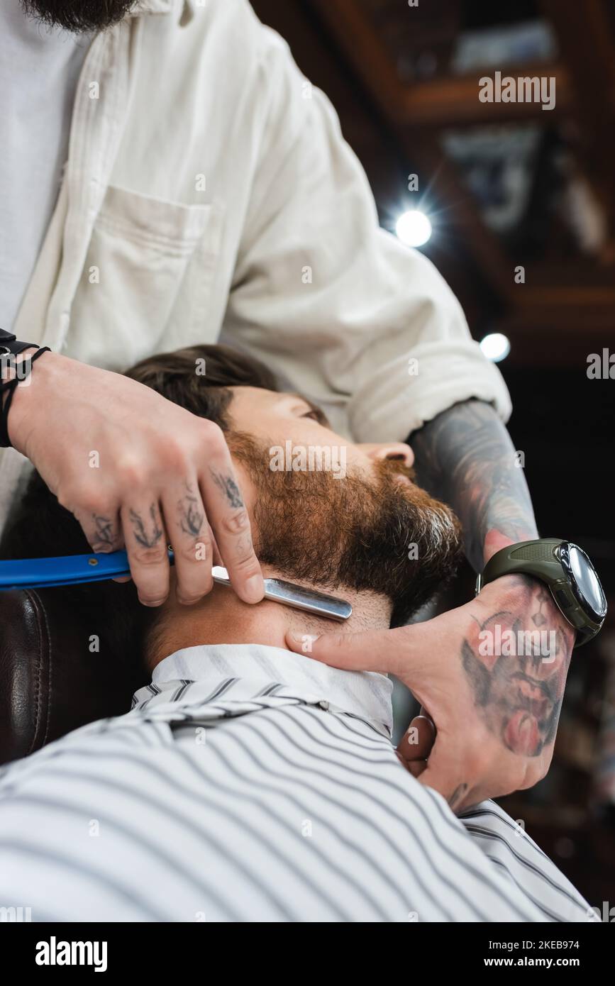 tattooed hairdresser shaving neck of bearded man with straight razor ...