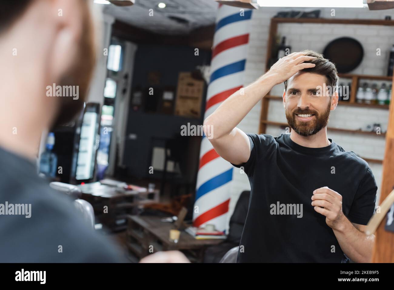 brunette bearded man smiling while looking in mirror and touching hair ...