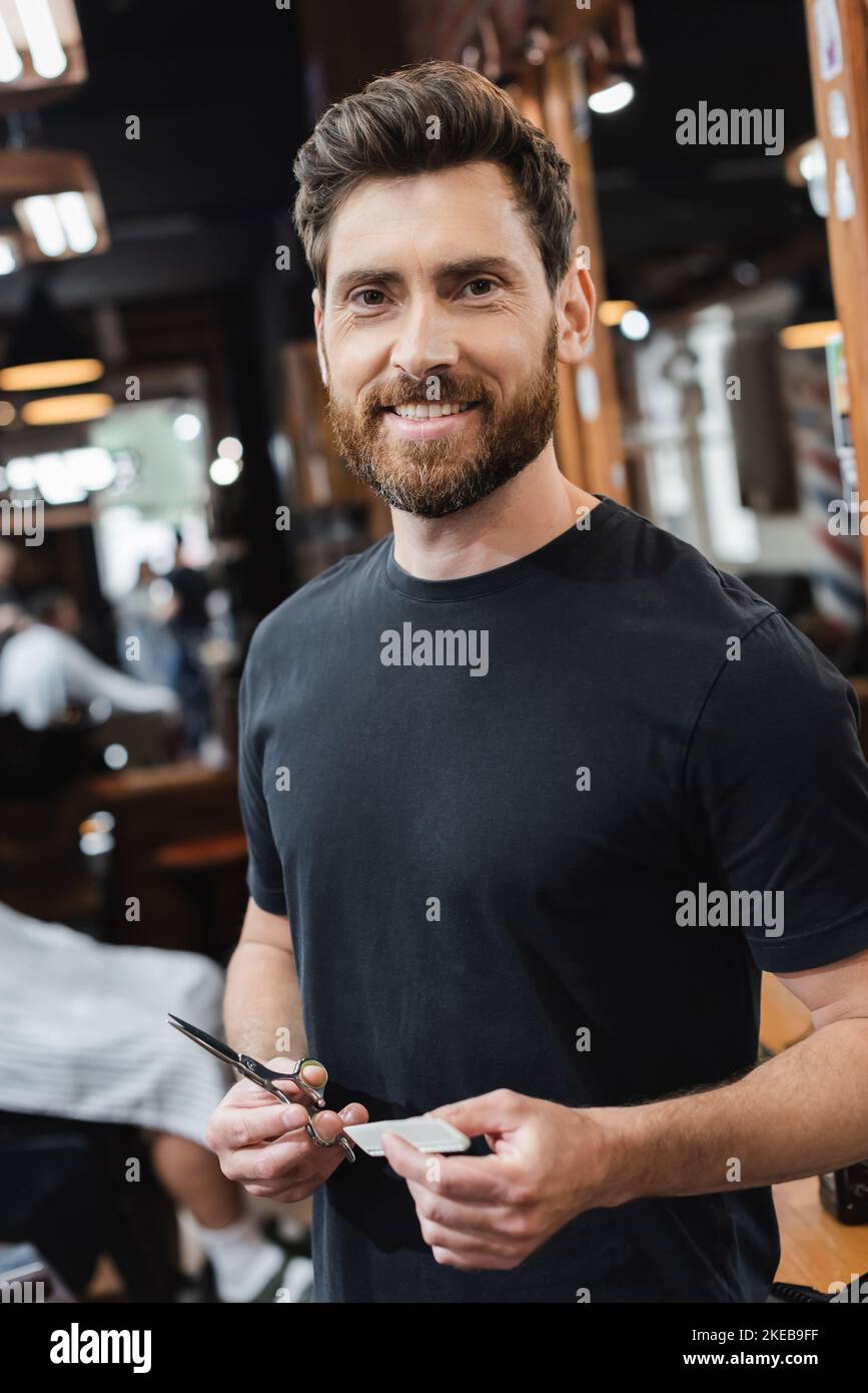 happy bearded barber holding scissors and comb while looking at camera ...