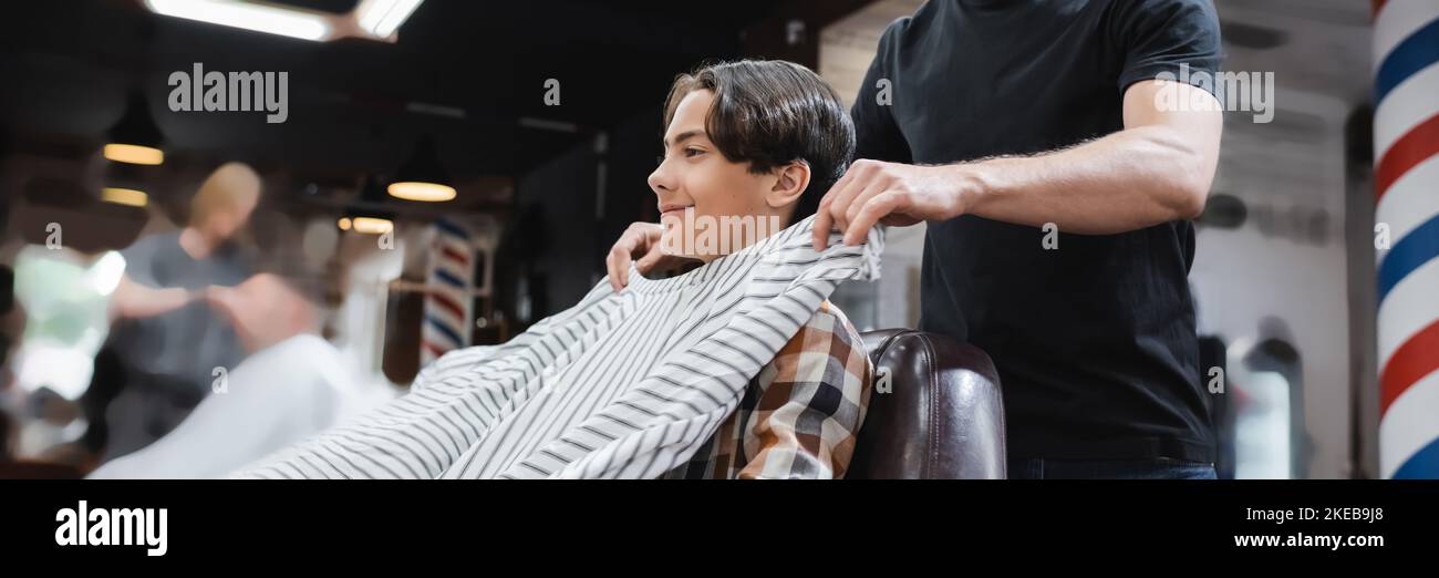 side view of barber putting hairdressing cape on smiling teen boy in ...
