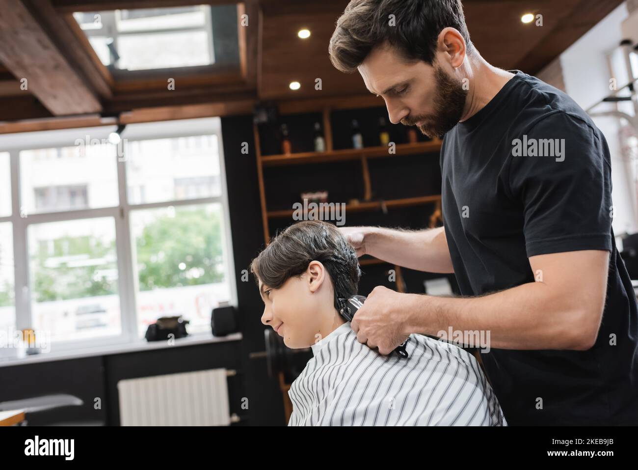 side view of bearded barber trimming hair of teen boy with electric ...