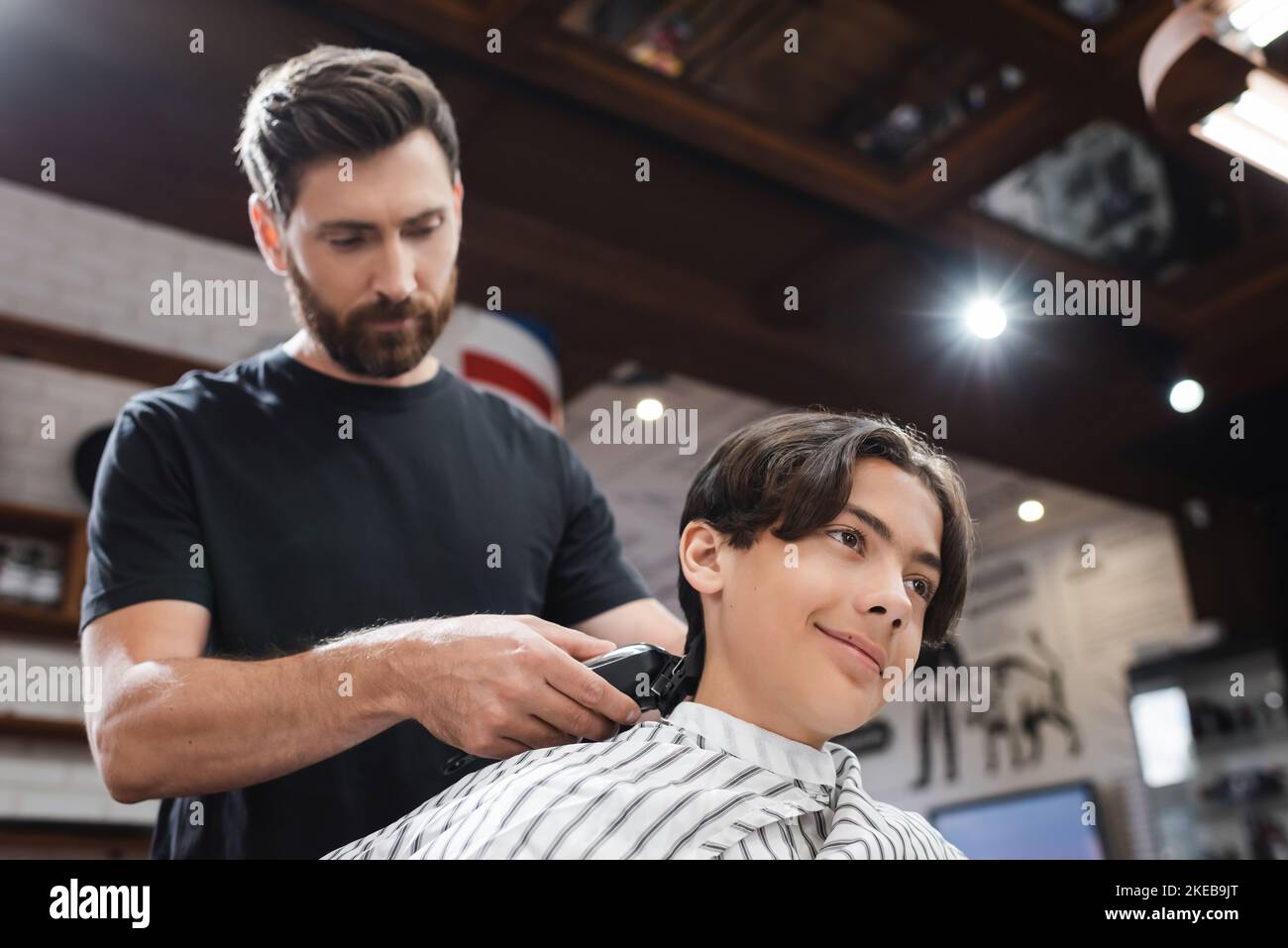 teen boy in hairdressing cape smiling while barber trimming his hair in ...