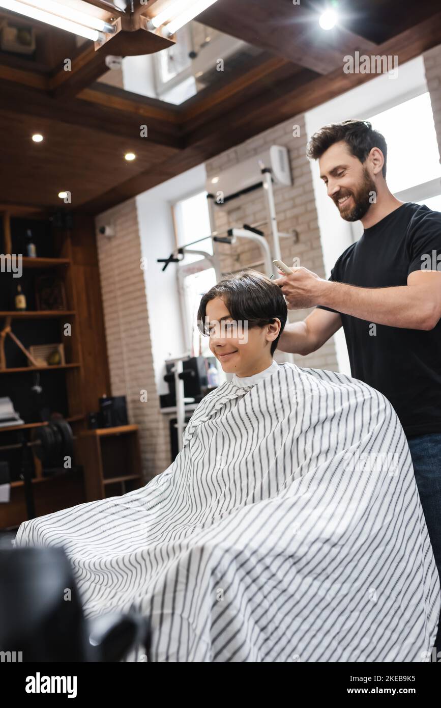 bearded barber working with teenage client sitting in hairdressing cape ...