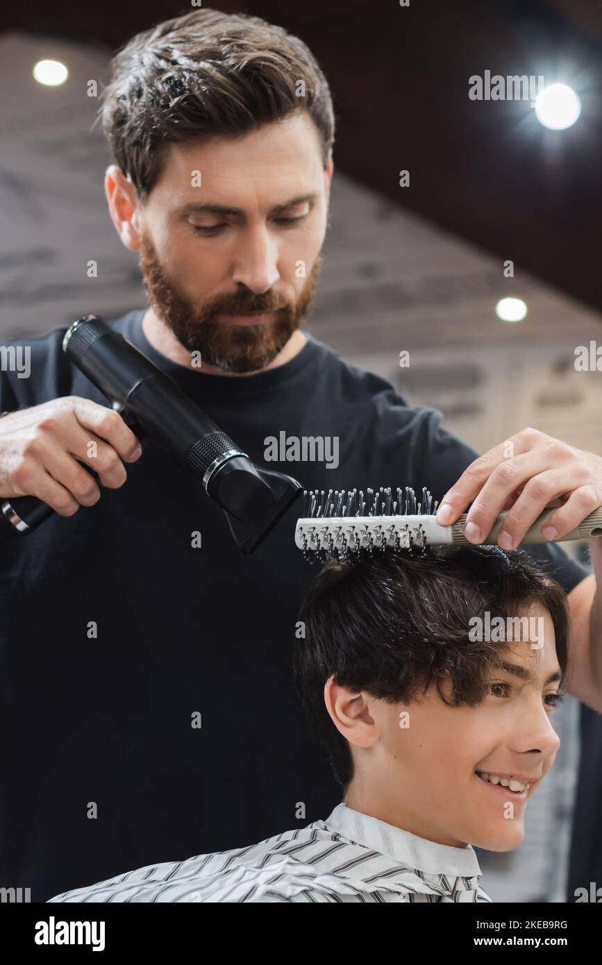 Barber drying and combing hair of smiling teen boy in beauty salon ...