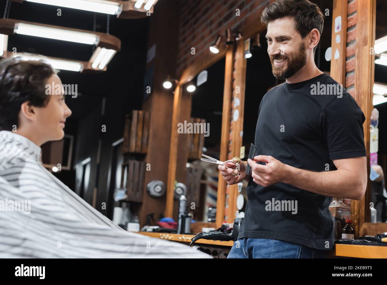Smiling barber holding comb and scissors near blurred teenager in ...
