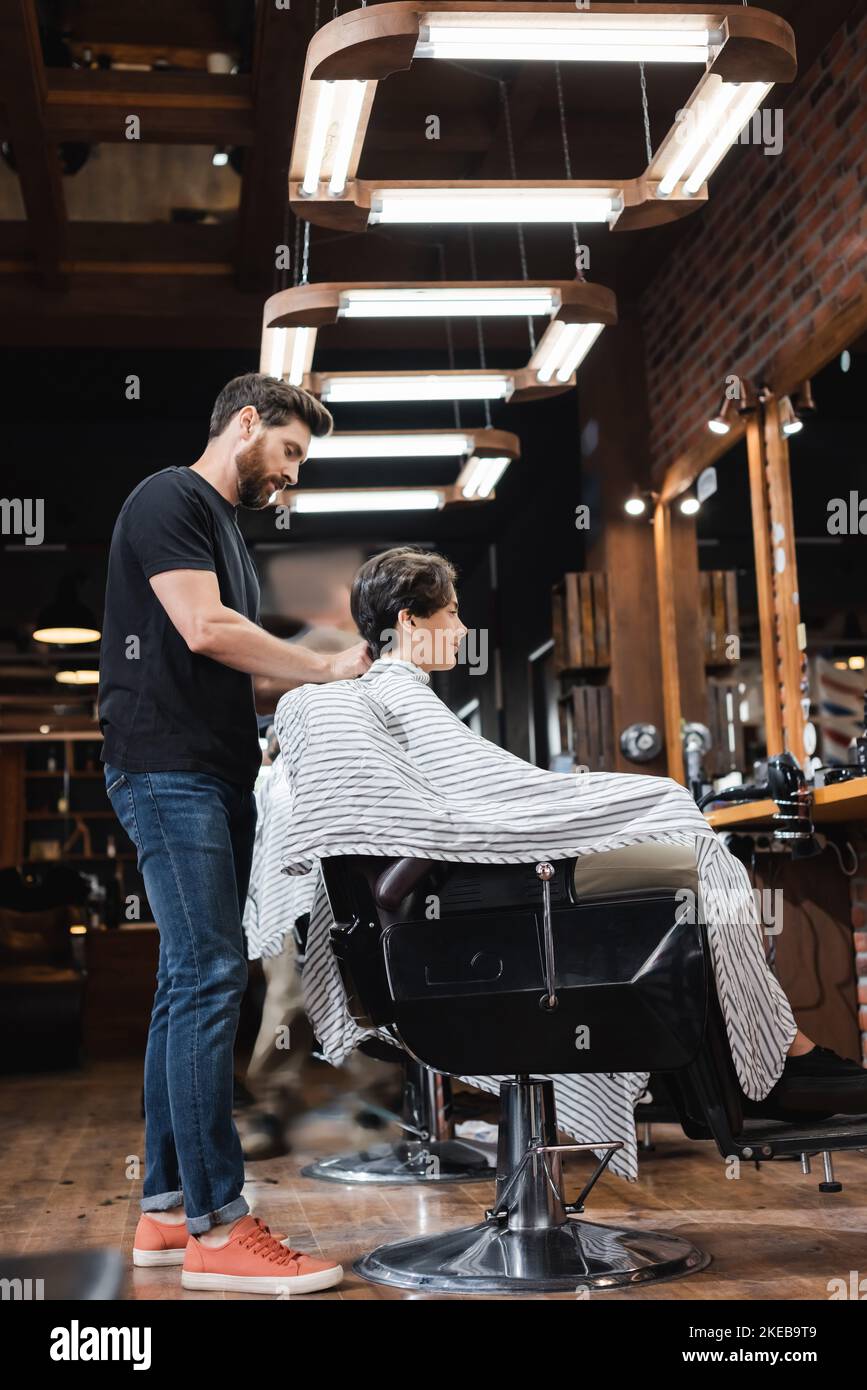 Side view of bearded hairstylist wearing cape on teenage client in ...