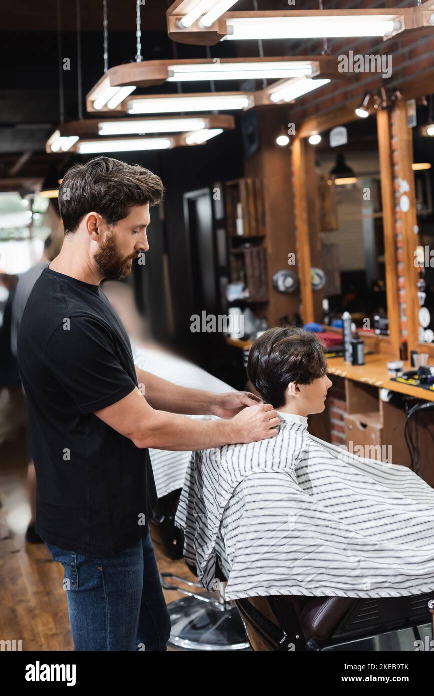 Side view of barber wearing hairdressing cape on teen boy in barbershop ...