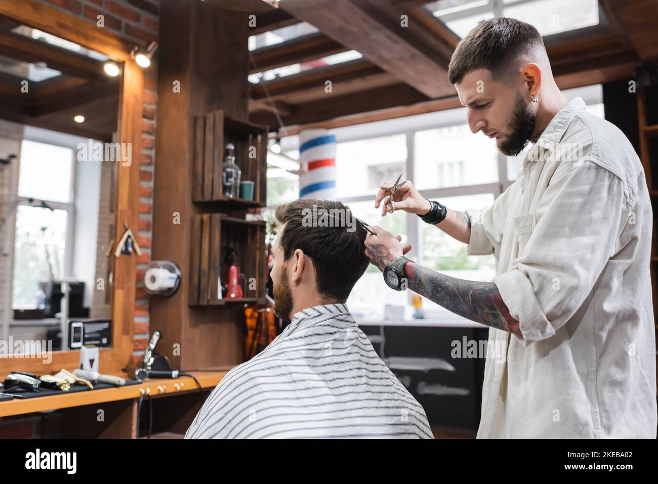 Tattooed barber combing hair of client and holding scissors in ...